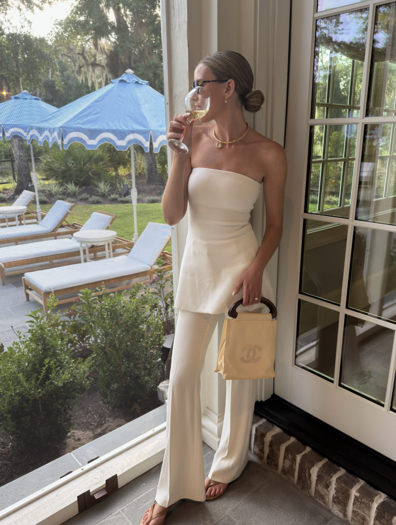 Woman in elegant white strapless top and wide-leg pants holding beige Chanel handbag and glass of white wine by patio door